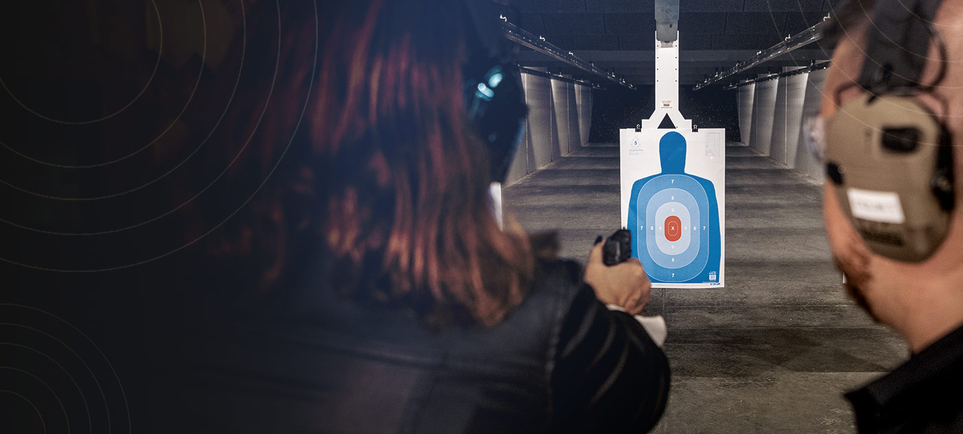 Two people back toward cameras, facing down range, one woman aiming gun ...